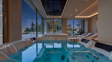 Piscina interior moderna com cobertura de vidro, rodeada por espreguiçadeiras e vista para o exterior.