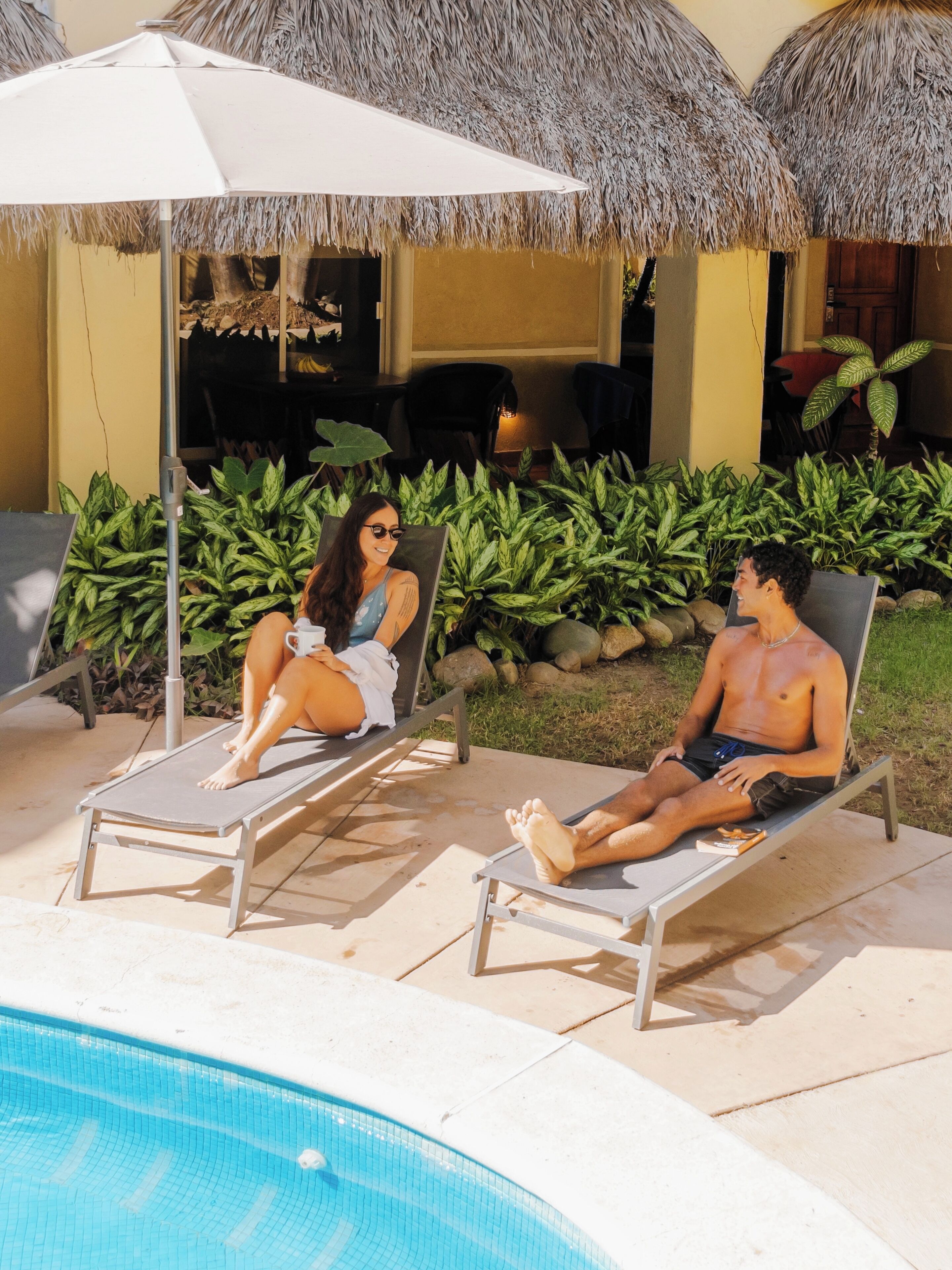 Duas espreguiçadeiras à beira da piscina, com uma estrutura de telhado de palha e plantas em vasos ao fundo.