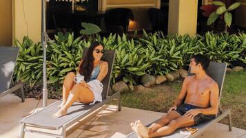 Duas espreguiçadeiras à beira da piscina, com uma estrutura de telhado de palha e plantas em vasos ao fundo.