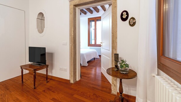 Um corredor com chão de madeira, uma televisão sobre uma mesa, uma porta que dá para um quarto e uma janela com cortinas.