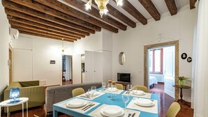Uma sala de jantar com uma mesa azul posta para quatro pessoas, vigas de madeira no teto e uma televisão ao fundo.