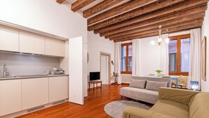 Cozinha moderna com armários brancos e vigas de madeira, sala de estar com sofá bege e área de jantar com mesa e cadeiras.