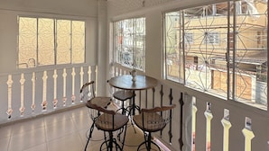 Quarto com varanda, mesa redonda e quatro cadeiras, e janelas amplas com grades de metal decorativas.