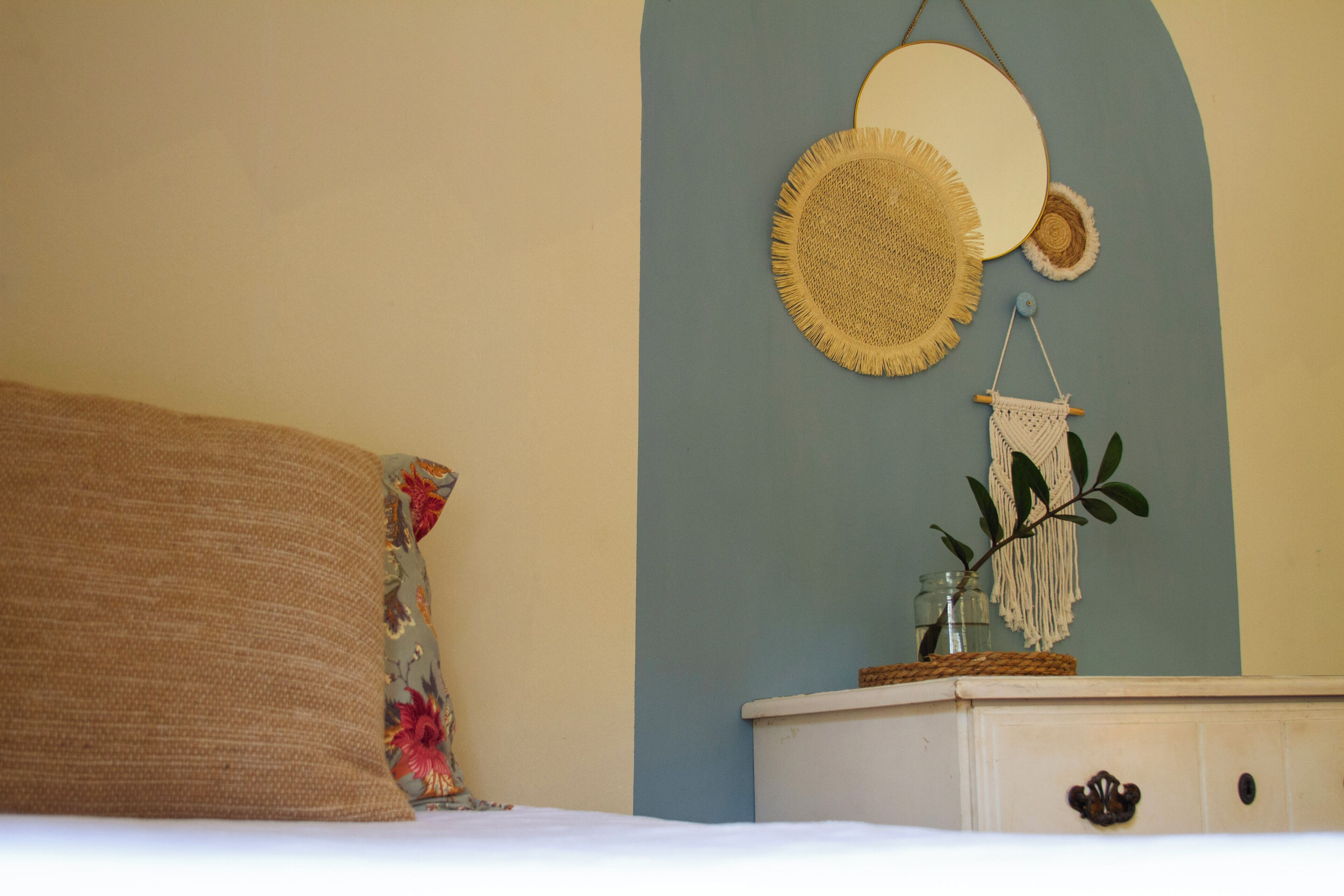 Quarto com cama, penteadeira e detalhes decorativos nas paredes, incluindo cestos de vime e um detalhe em macramé.