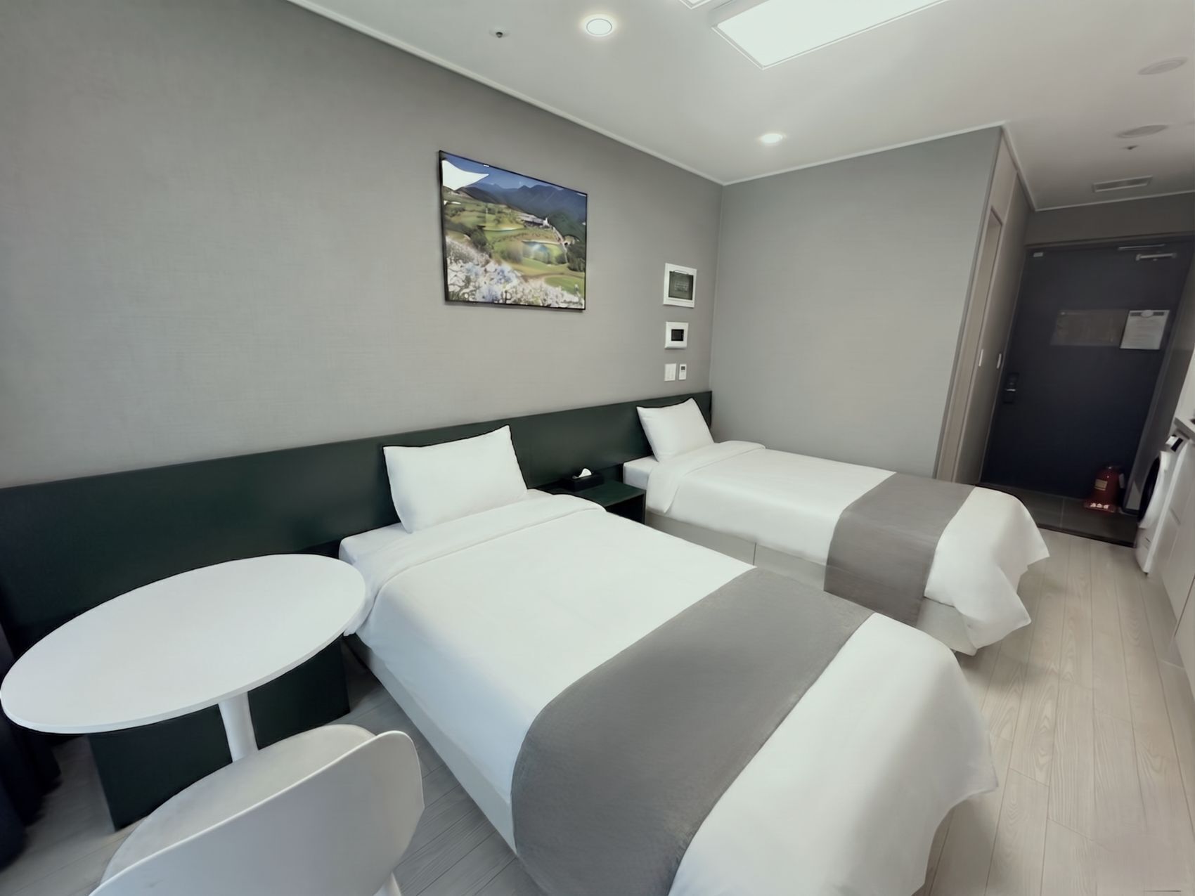 Quarto de hotel com duas camas, uma mesinha e uma cadeira. Há um quadro na parede e uma porta à direita.