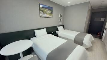 Quarto de hotel com duas camas, uma mesinha e uma cadeira. Há um quadro na parede e uma porta à direita.