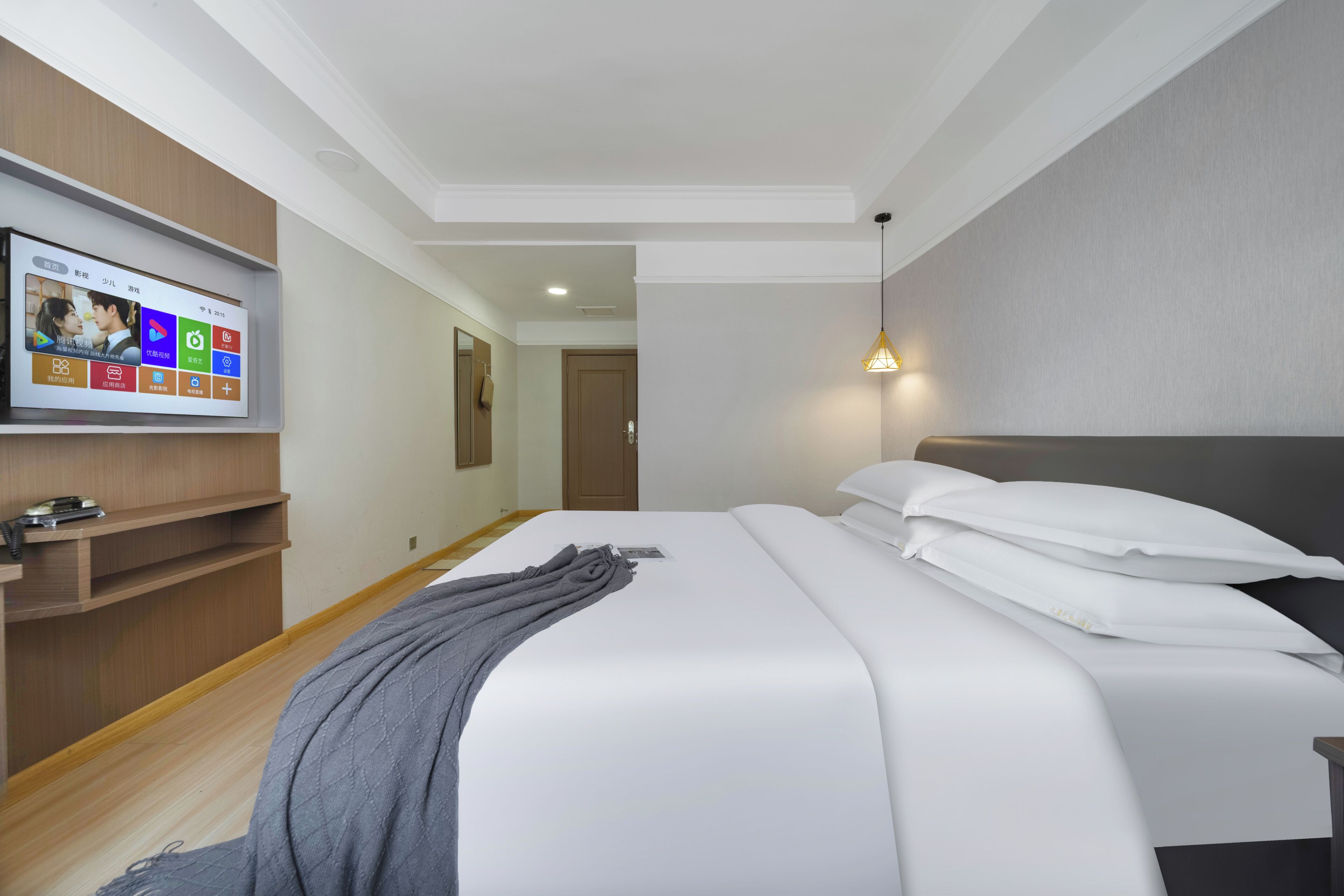 Quarto de hotel com cama, televisão fixada na parede, prateleira de madeira e porta que dá para outro quarto.