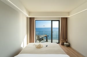 Quarto de hotel com uma cama grande, vista para o mar, varanda com mesa e cadeira, e um vaso com flores.