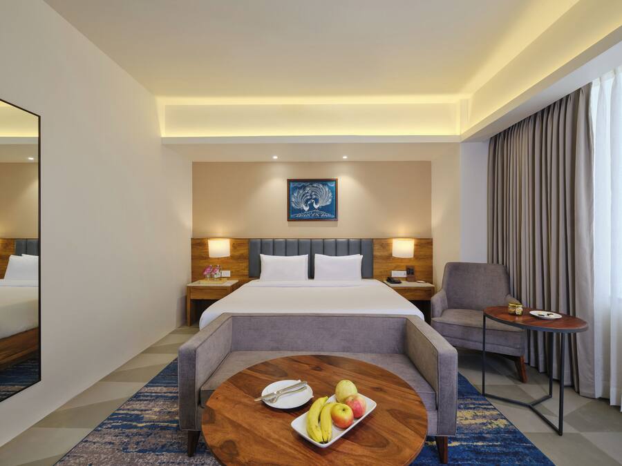 Quarto de hotel com cama, mesas de cabeceira, uma mesa de madeira redonda com frutas, uma poltrona cinza e uma grande janela com cortinas.