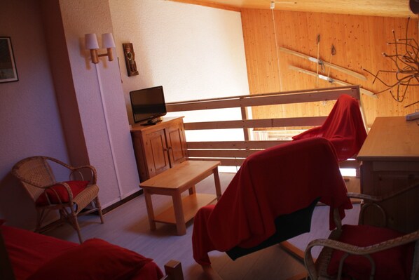 Um quarto com um banco de madeira, cadeiras de vime, uma televisão sobre um armário de madeira e um cobertor vermelho jogado sobre uma cadeira.
