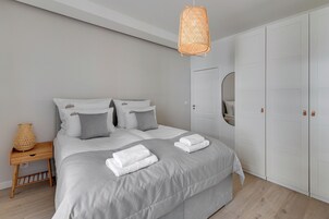 Quarto moderno com cama grande, roupa de cama cinzenta, mesa de cabeceira de madeira, luminária trançada e guarda-roupa.