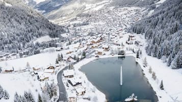 Uma aldeia coberta de neve, com construções agrupadas em torno de um lago central, rodeada por montanhas florestadas.