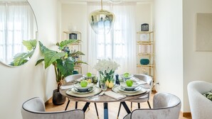 Uma área de refeições com uma mesa redonda posta para quatro pessoas, rodeada por cadeiras. Há plantas em vasos, um vaso com flores e um candelabro acima.