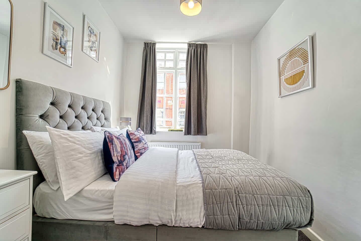 Quarto com cabeceira estofada, cama com roupa de cama estampada, mesa de cabeceira e janela com cortinas.