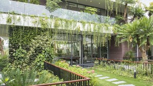 Um edifício moderno com fachada de vidro, jardins verticais exuberantes e área externa bem cuidada, com um caminho e assentos.
