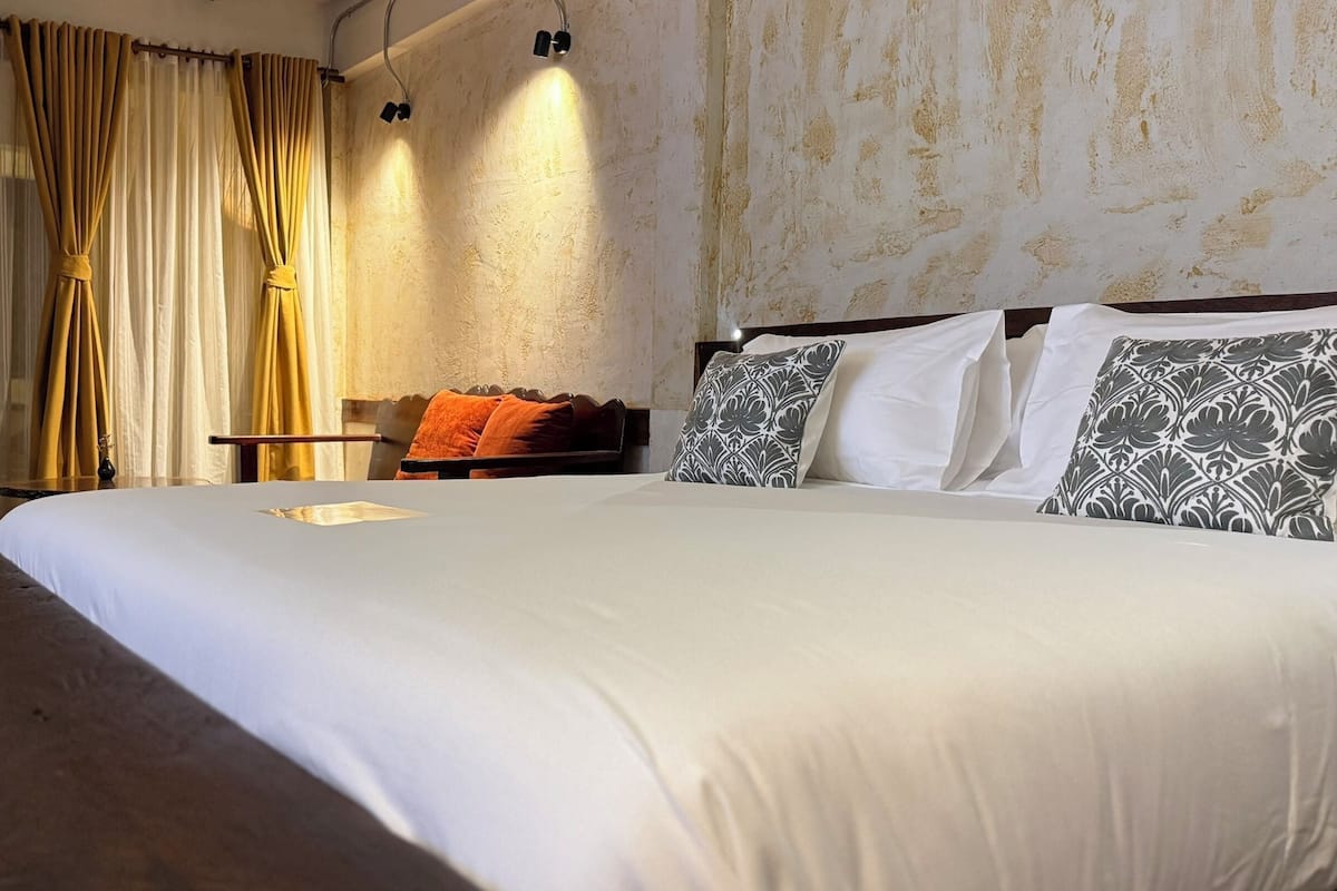 Cama bem arrumada com roupa de cama branca, cabeceira com almofadas estampadas e mesa de cabeceira de madeira com uma almofada vermelha.