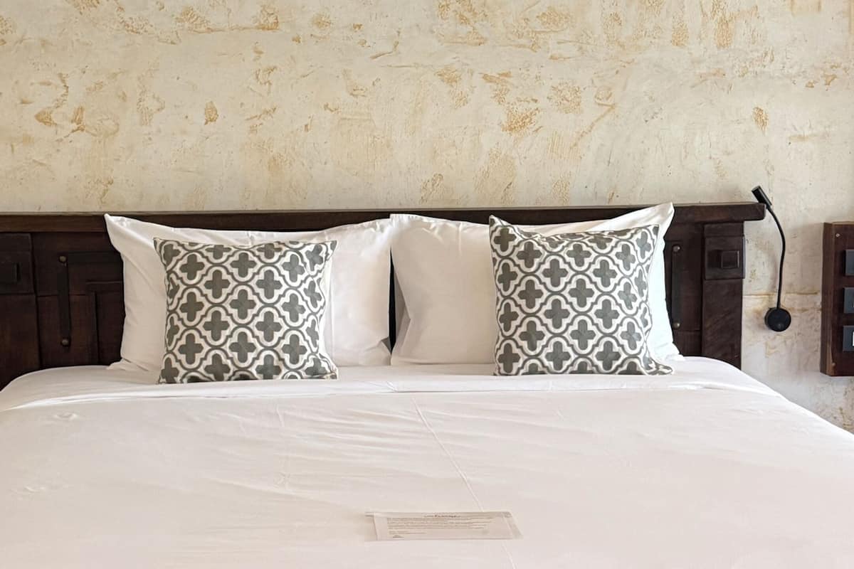 Uma cama com lençol branco, duas almofadas estampadas e cabeceira de madeira.