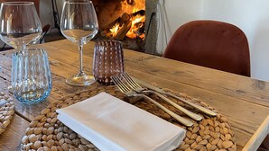 Mesa posta com taças de vinho, talheres e um jogo americano de tecido. Uma lareira é visível ao fundo.