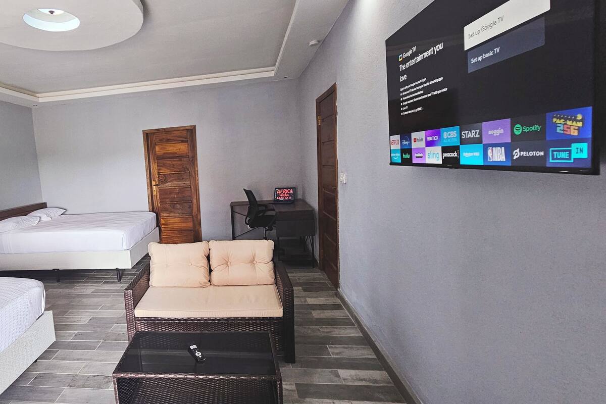 Quarto de hotel com duas camas, um sofá pequeno, uma TV e uma luminária no teto.