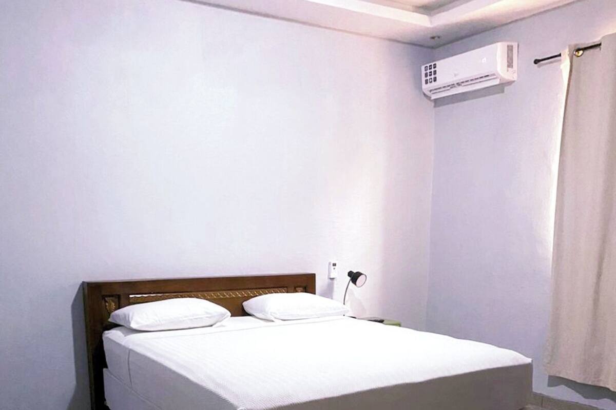 Quarto individual com uma cama e cabeceira de madeira, roupa de cama branca, abajur e ar condicionado na parede.