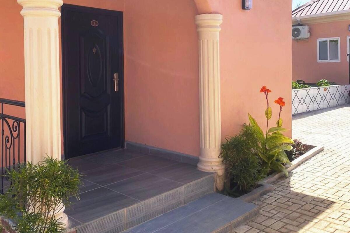 Um edifício com fachada rosa, porta preta e colunas brancas. Há degraus levando à entrada, e um pequeno jardim com plantas e flores.