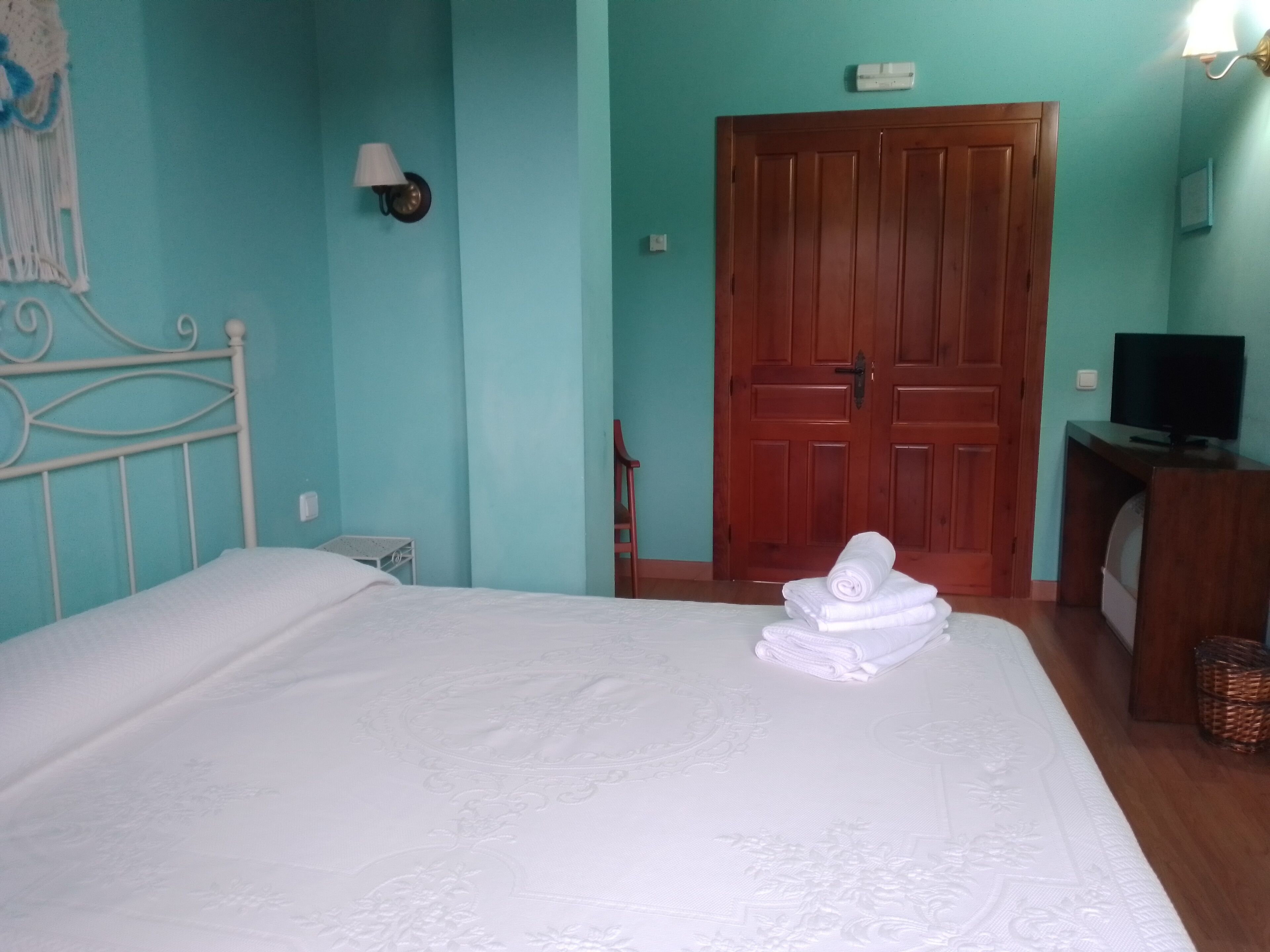 Quarto com cama, porta de madeira, televisão e luminária fixa na parede.