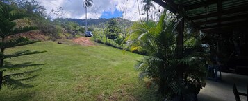 Área gramada com uma trilha de terra que leva a um veículo branco, cercada por vegetação exuberante e uma montanha ao fundo.