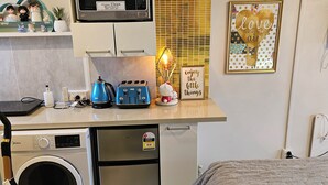 Uma cozinha compacta com micro-ondas, torradeira e máquina de lavar roupa. Há uma cama com um cobertor cinzento e uma parede com um letreiro 'LOVE' e uma frase emoldurada.