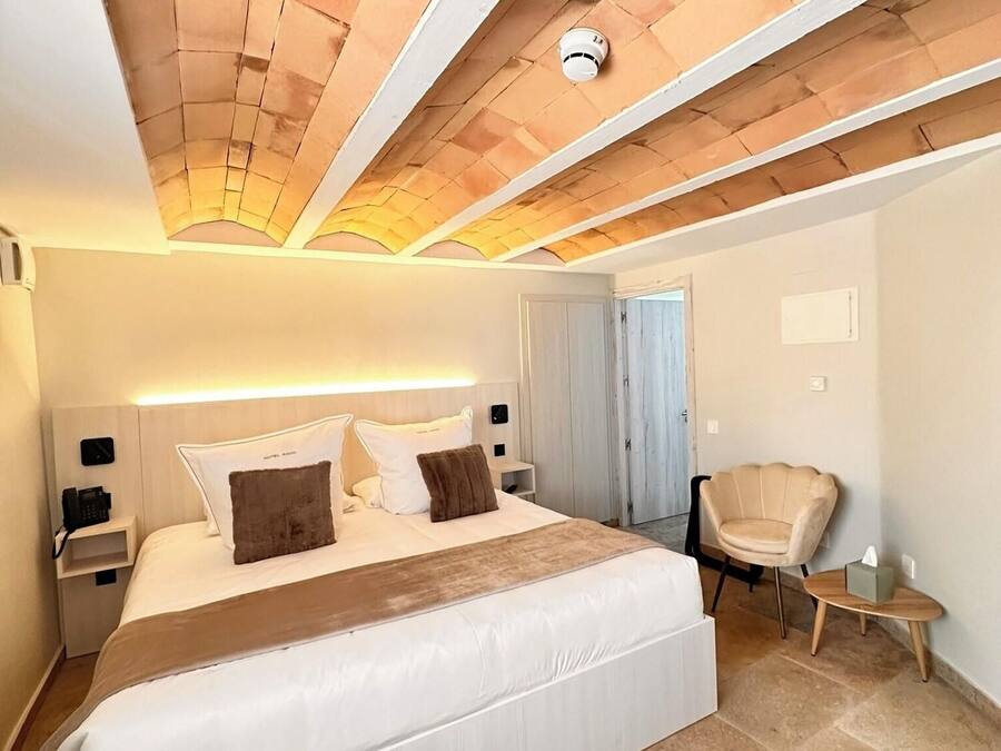 Quarto de hotel com uma cama grande, mesa de cabeceira com telefone, uma cadeira e uma mesinha com caixa de lenços.