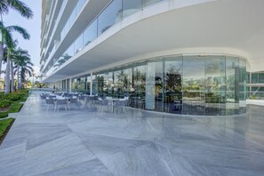 Exterior moderno de hotel com fachada de vidro, área de estar ao ar livre e palmeiras.