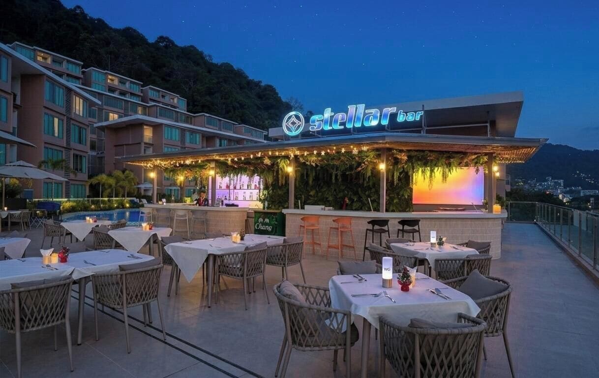 Uma área de refeições ao ar livre com mesas, cadeiras, um bar e uma piscina ao fundo.