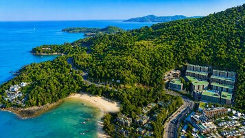 Vista aérea de uma área costeira com praia, floresta densa e um conjunto de edifícios com telhado verde.