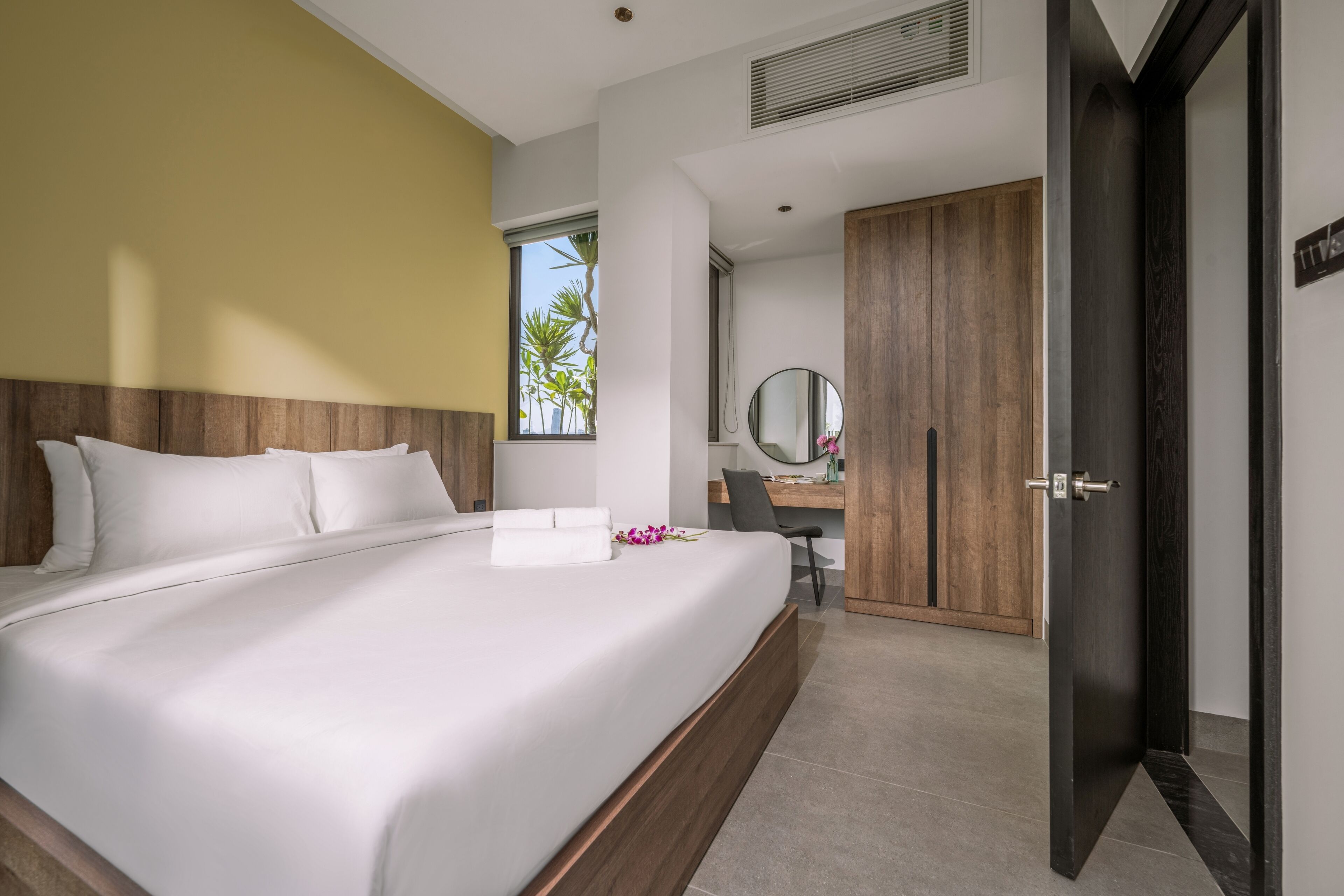 Quarto de hotel moderno com uma cama grande, um guarda-roupa de madeira, uma mesa de escritório com espelho e vista para palmeiras pela janela.
