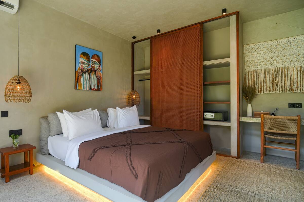 Quarto com cama, mesas de cabeceira, um quadro, um separador tecido e uma cadeira.