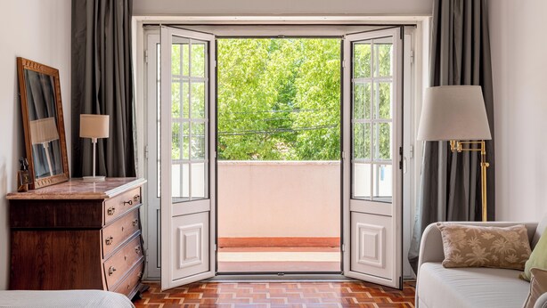 Quarto com penteadeira de madeira, cama, mesa de cabeceira, abajur e vista para uma varanda com vegetação.