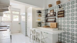 Uma cozinha moderna com armários brancos, bancada branca e painel de fundo estampado. Há duas cadeiras brancas, uma mesa pequena e uma cesta no chão.