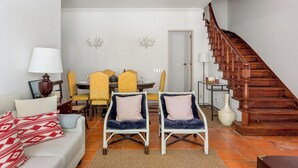 Sala de estar com sofá, duas poltronas, mesa de jantar com cadeiras amarelas, escada e porta.
