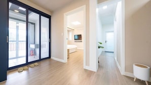 Um apartamento moderno com uma grande porta de correr de vidro que dá para uma varanda, um quarto com cama e televisão, e um banheiro pequeno com chuveiro.