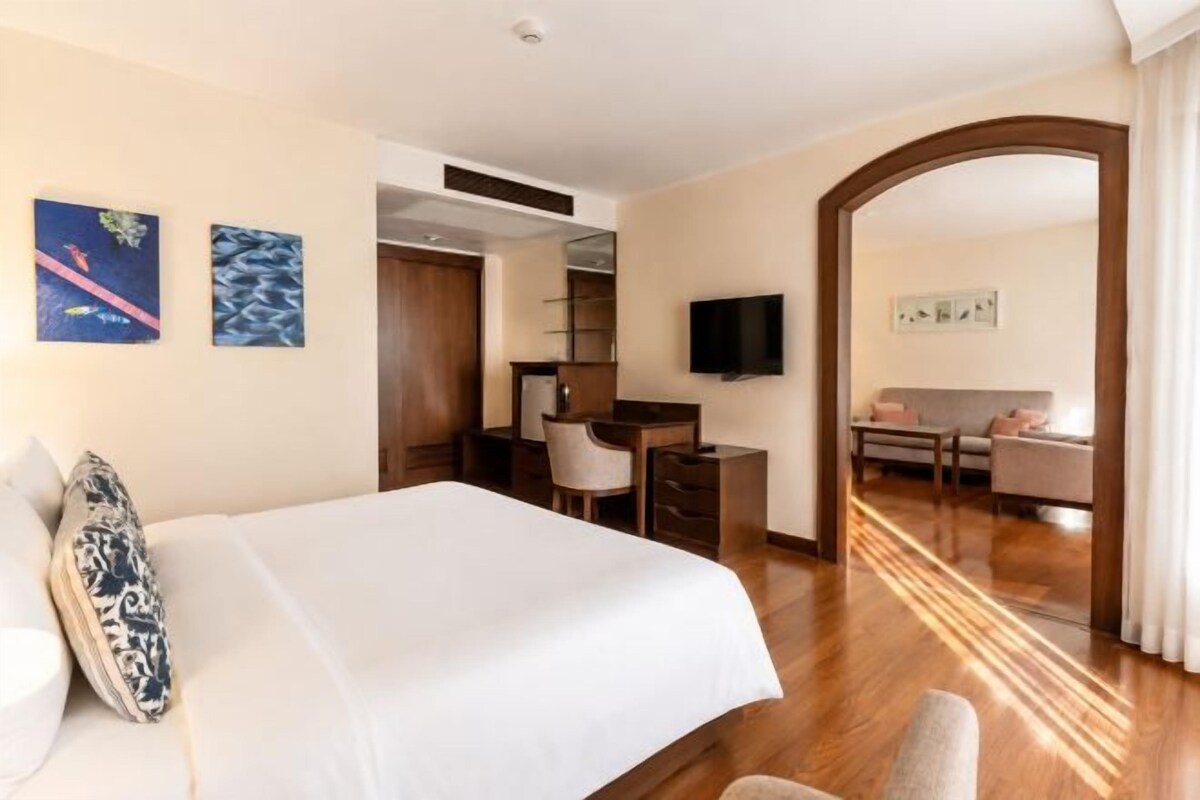 Quarto de hotel com cama, escrivaninha, cadeira, televisão e vista para outro quarto através de um arco.