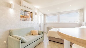 Quarto de hotel moderno com sofá de cor clara, cama com cabeceira de madeira, uma mesinha e uma janela grande com vista.
