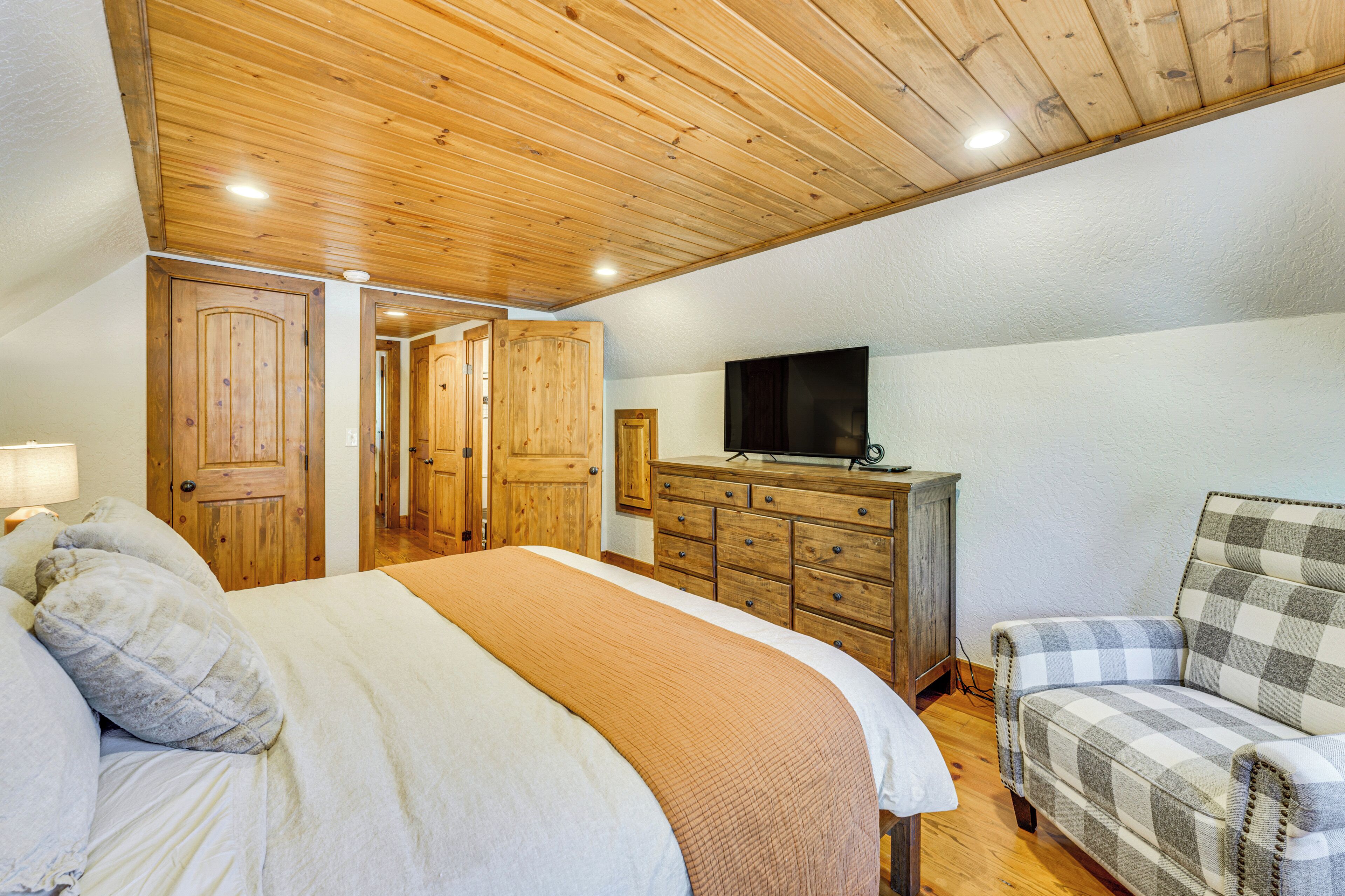 Quarto com teto de madeira, cama com manta bege, cômoda de madeira, televisão em cima e poltrona xadrez.