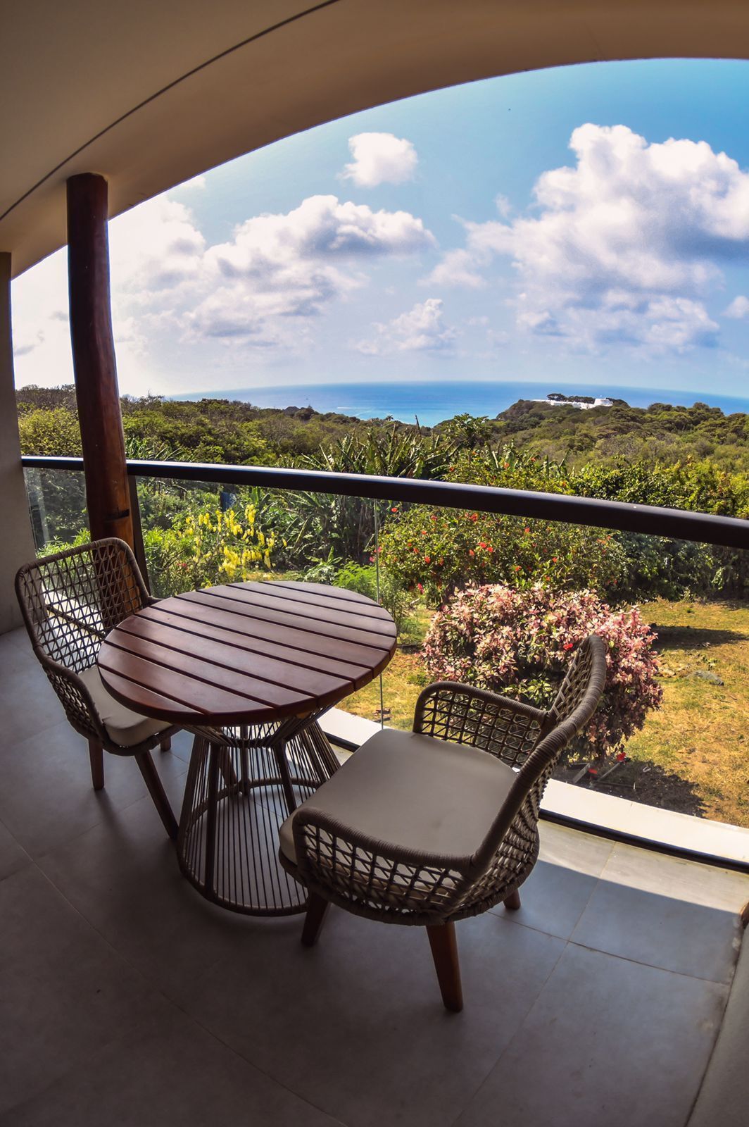 Uma varanda com uma mesa redonda e duas cadeiras de vime, com vista para uma paisagem exuberante com árvores e o oceano ao longe.
