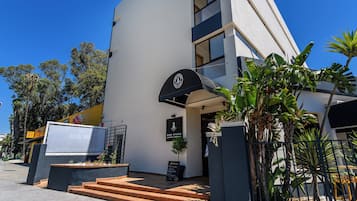 Um edifício moderno com fachada curva, toldo preto e placa indicando ser um hotel. Há degraus levando à entrada, e um vaso de plantas com uma pequena árvore.