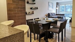 Uma área de refeições moderna com mesa e cadeiras de vidro, bancada de cozinha e sala de estar com sofá e mesa de centro.