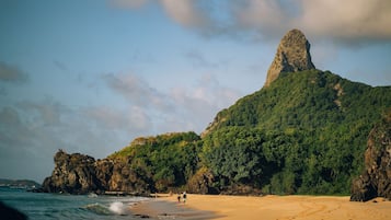 Uma praia com um pico rochoso imponente ao fundo, céu claro e pegadas na areia.