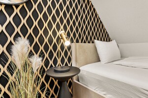 Quarto moderno com parede estampada em padrão geométrico, mesa de cabeceira com abajur e cama com roupa de cama branca.