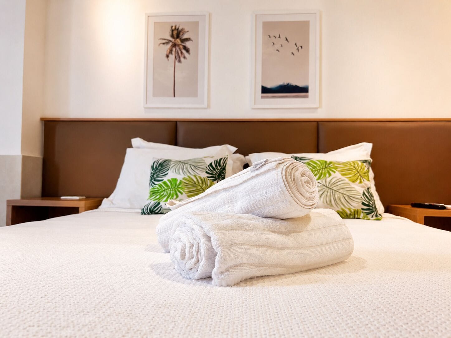 Cama bem arrumada com roupa de cama branca, cabeceira marrom e almofadas com tema tropical.