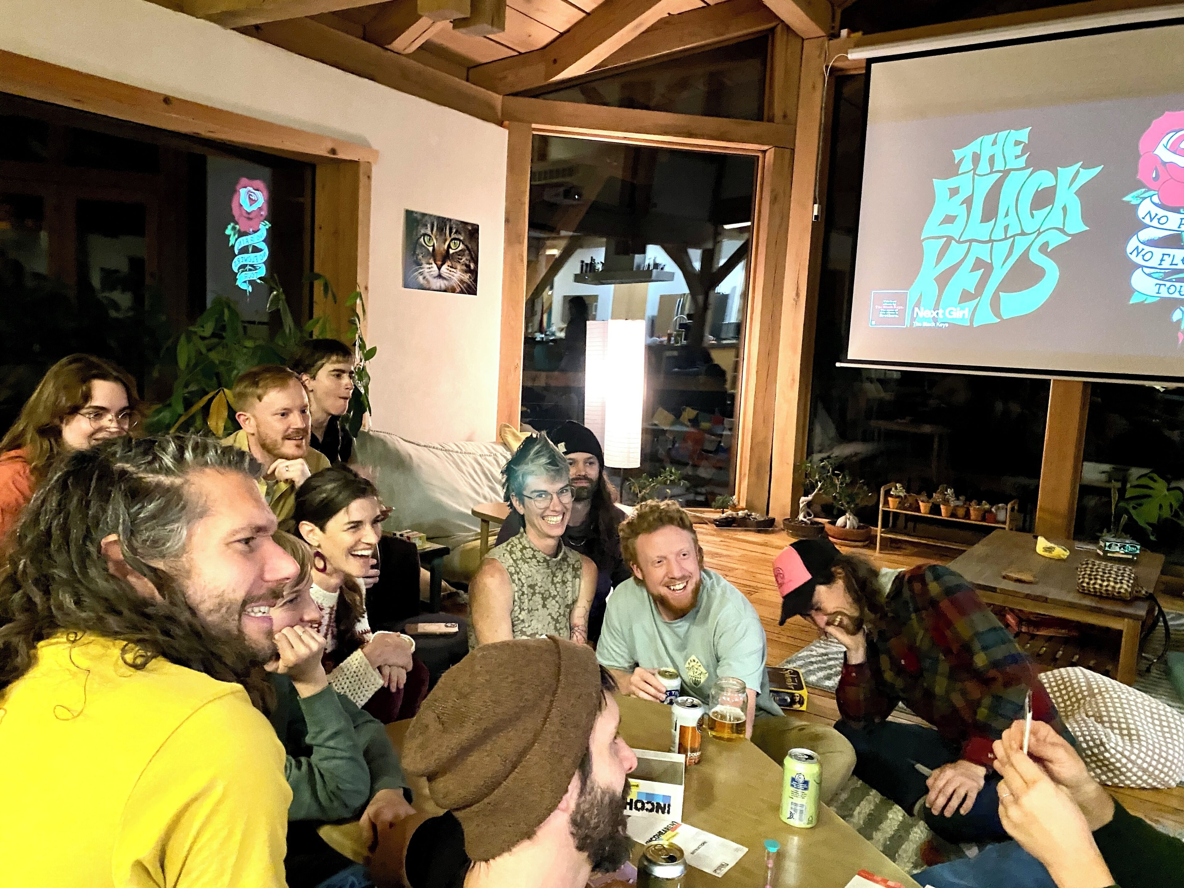 Um grupo de pessoas sentado à volta de uma mesa numa sala com vigas de madeira no teto, um ecrã de projeção a exibir o logótipo de 'The Black Keys', e vários vasos com plantas.