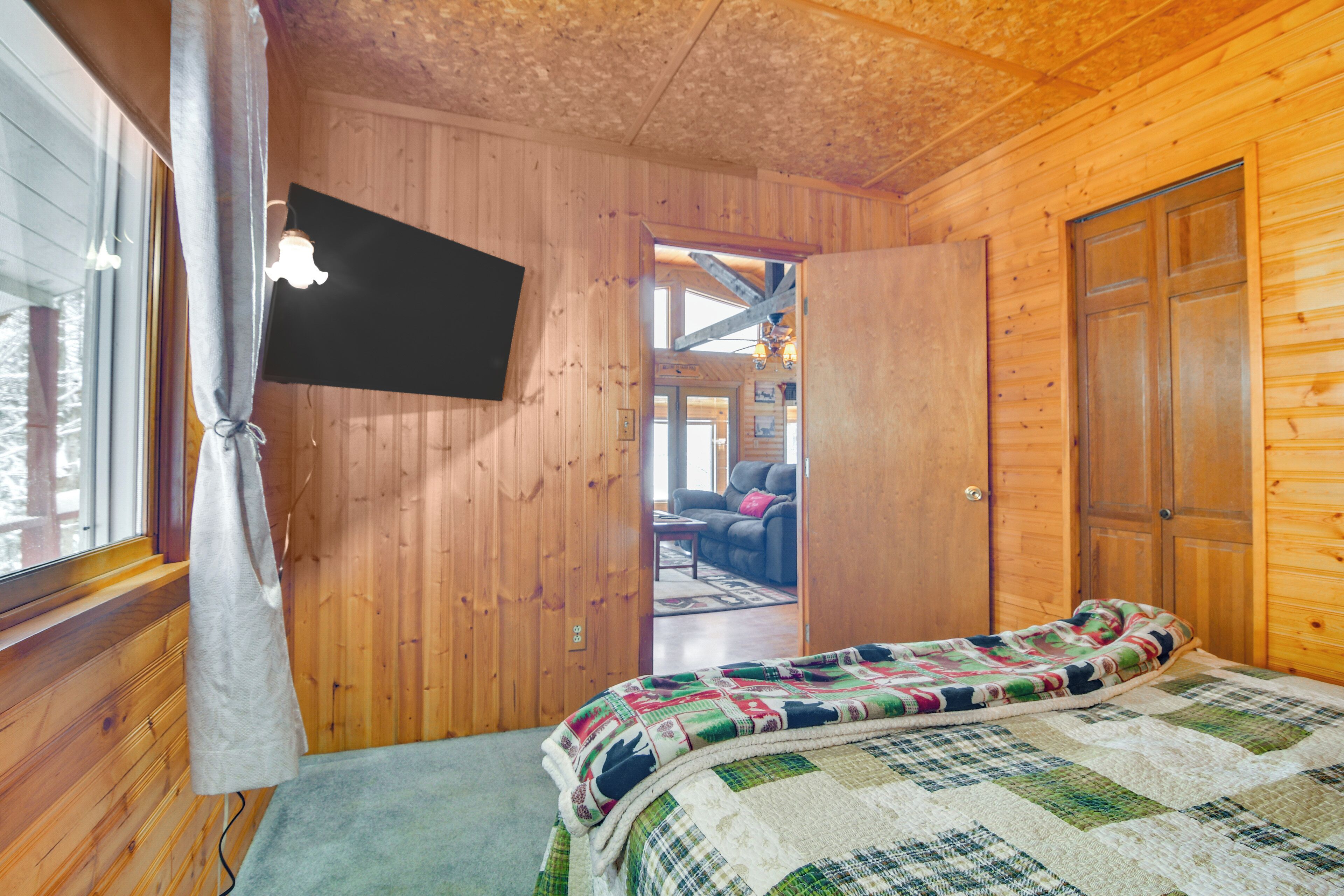 Quarto de cabana de madeira com uma cama, uma televisão fixada na parede e uma porta que dá para outro quarto.