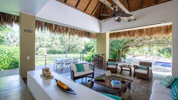 Uma sala de estar ampla, com teto alto, ventilador de teto e vista para uma piscina e vegetação exuberante.